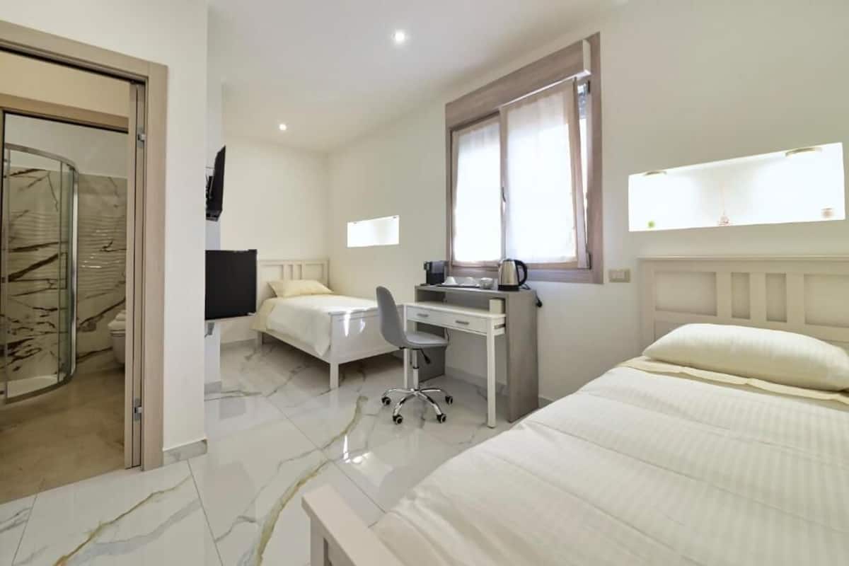 Quarto de hotel com cama, escrivaninha, cadeira, televisão e área de chuveiro.