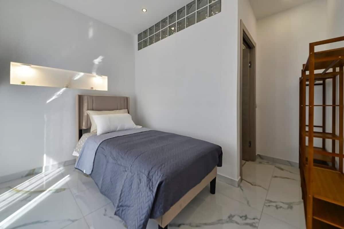 Quarto com cama, luminária na parede, porta e cadeira de madeira.