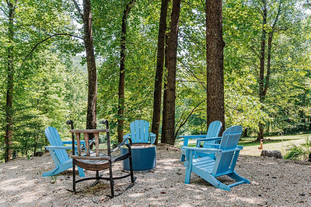 Área arborizada com área de estar externa, incluindo cadeiras Adirondack e uma mesa, cercada por árvores.