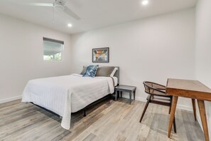 Quarto com cama, mesa pequena, cadeira, ventoinha no teto, janela e um quadro na parede.