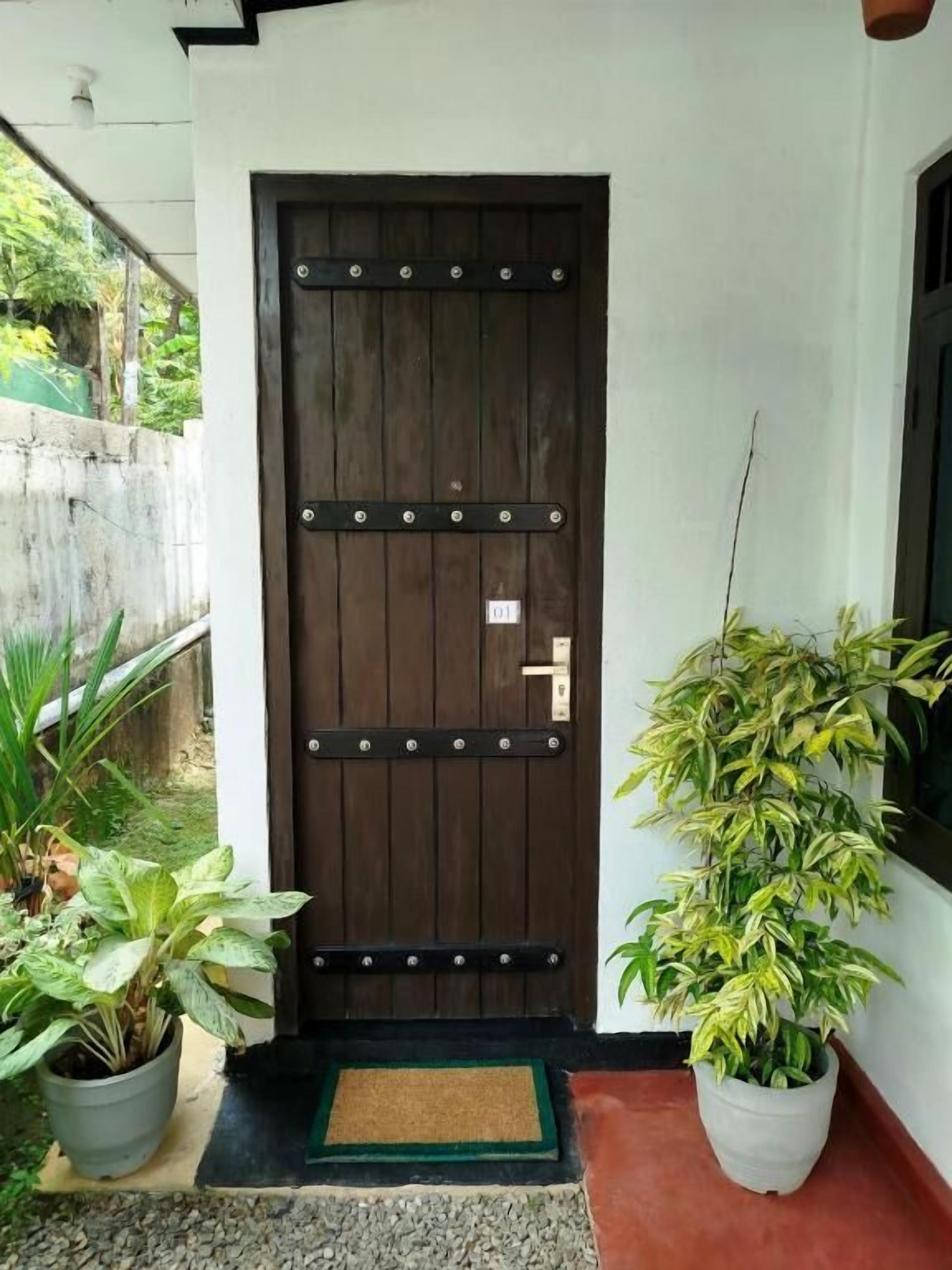 Uma porta marrom com detalhes metálicos, um tapete de entrada e plantas em vasos.
