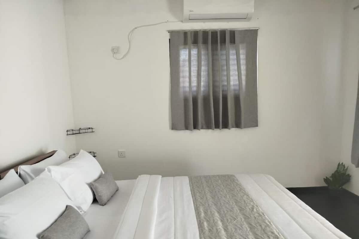 Quarto com cama, almofadas, mesa de cabeceira, ar condicionado fixado na parede e janela com cortinas.