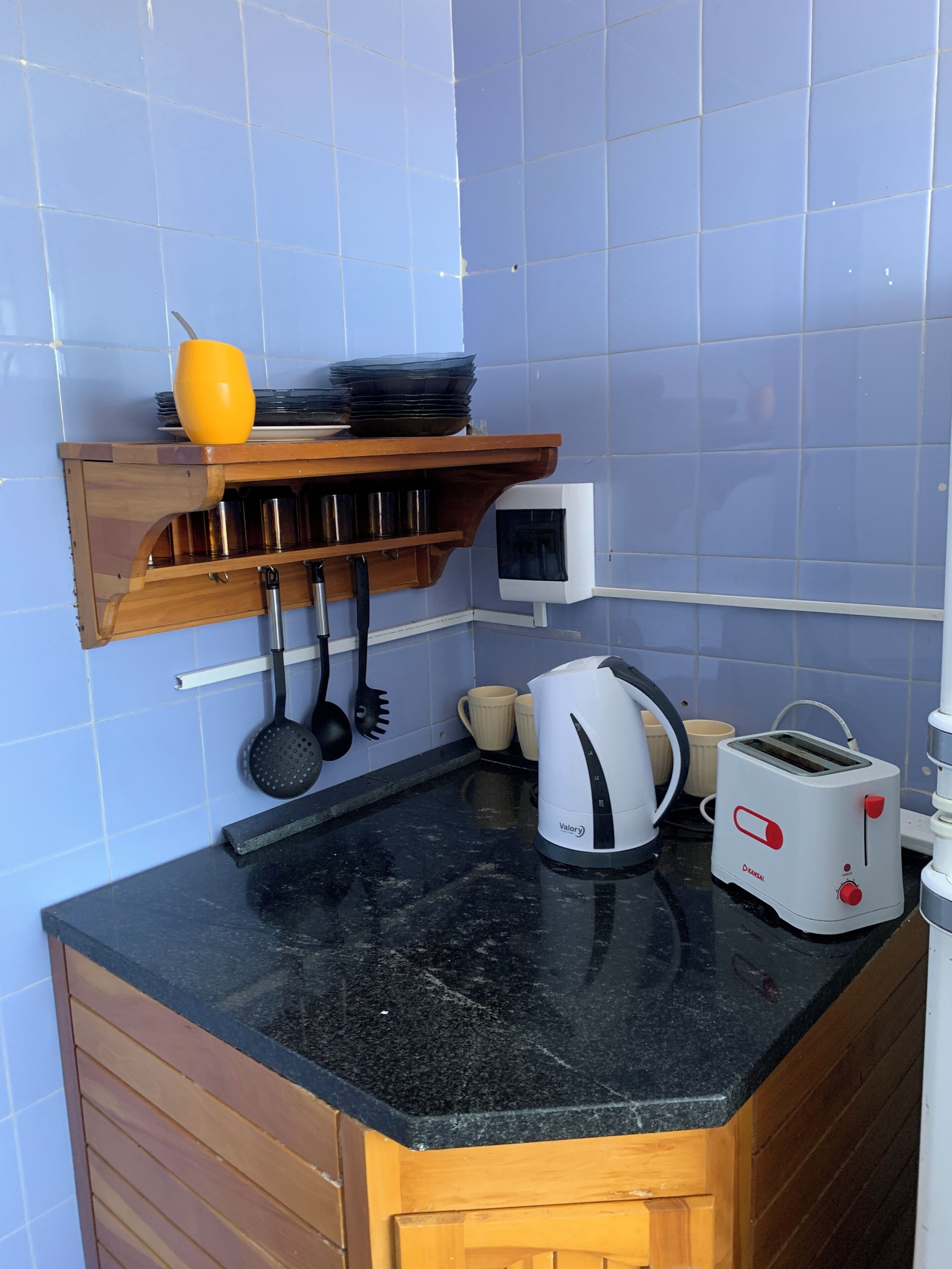 Cozinha com bancada preta, chaleira Valory, torradeira e prateleira com utensílios de cozinha.