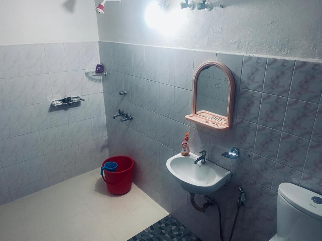 Casa de banho com lavatório, espelho e sanita. Há um balde vermelho no chão.
