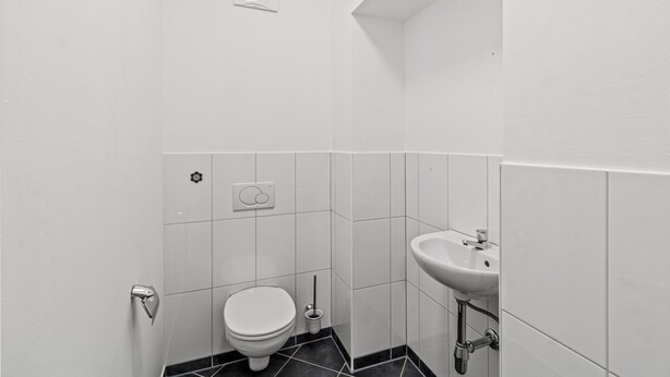 Uma casa de banho pequena e moderna, com sanita, lavatório e um candeeiro fixo na parede.