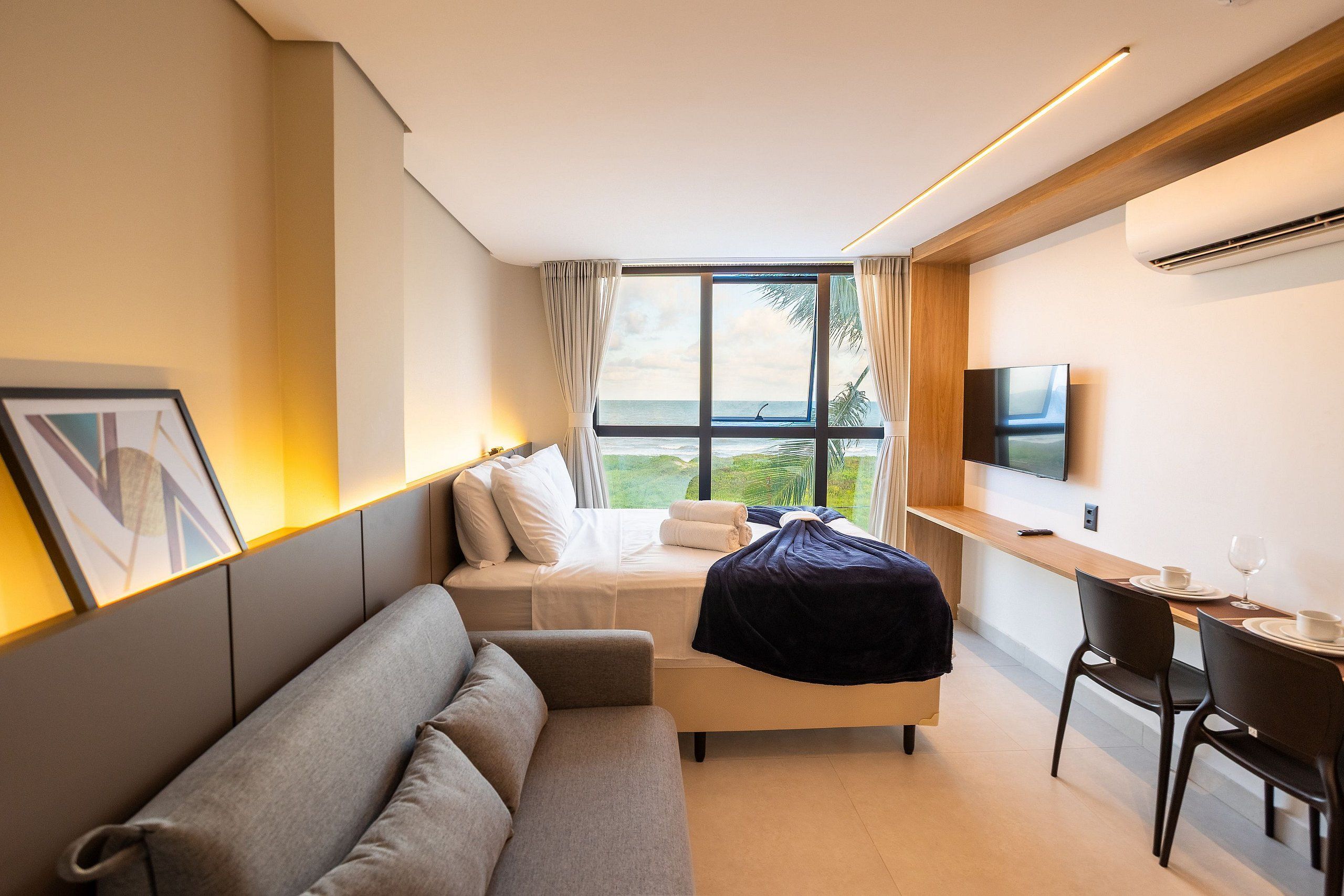 Quarto de hotel com cama, sofá, escrivaninha com cadeiras, televisão e janela com vista para o mar.