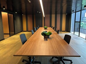 Uma sala de conferências com uma longa mesa de madeira, cadeiras pretas, plantas em vasos e amplas janelas.