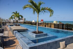 Uma piscina infinita no terraço, com palmeiras, espreguiçadeiras e vista para o oceano.