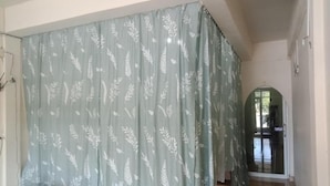 Quarto com uma grande janela, cortina com padrão de folhas, porta e piso ladrilhado.