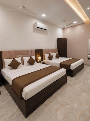 Quarto de hotel com duas camas, um ventilador de teto, unidade de ar condicionado, mesa de cabeceira com abajur e um guarda-roupa.