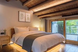 Quarto com teto de madeira, cama com roupa de cama branca, mesa de cabeceira, um vaso com flores e janela com vista para o verde.