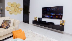 Uma sala de estar com sofá, almofadas laranja, TV fixada na parede exibindo diversos canais e um objeto decorativo na parede.