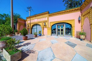 Um edifício com fachada rosa, grandes janelas em arco e um pátio ladrilhado com plantas em vasos.