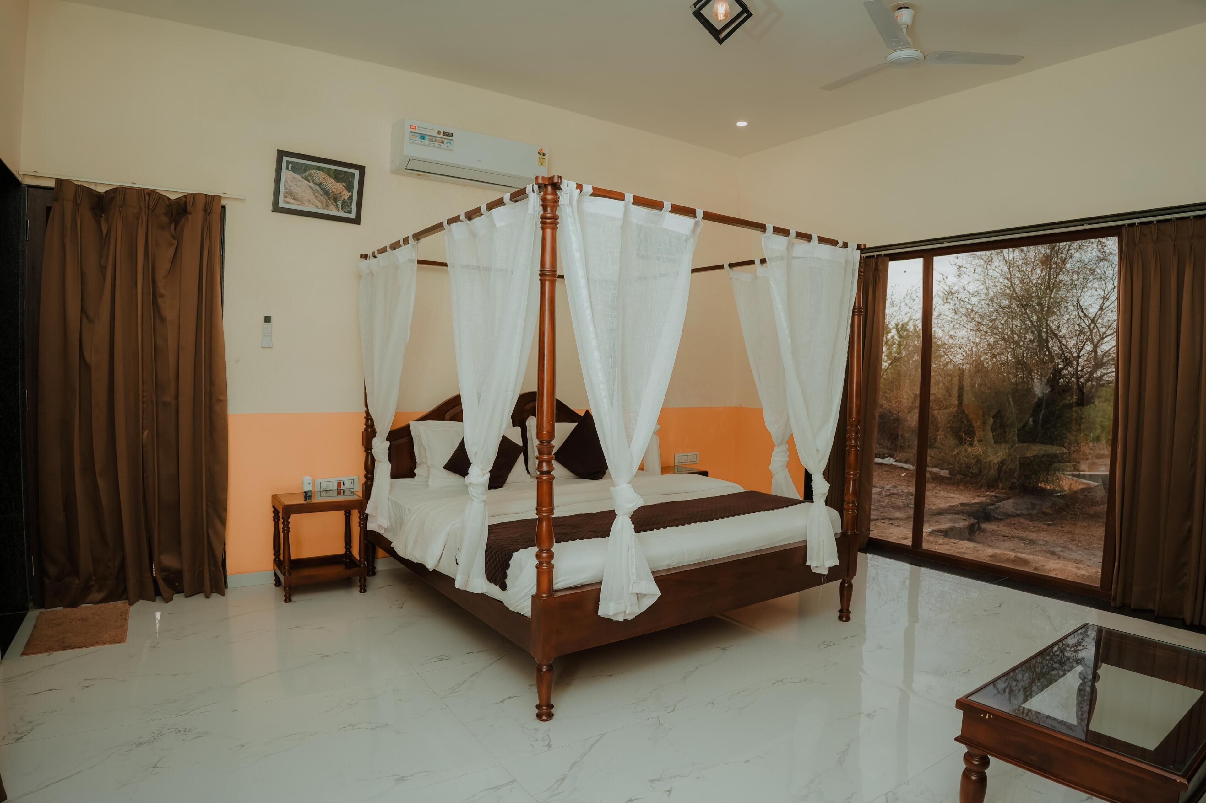 Uma cama de dossel com edredons brancos e almofadas castanhas, uma mesa de cabeceira, um ventilador de teto e uma janela grande com vista para árvores.