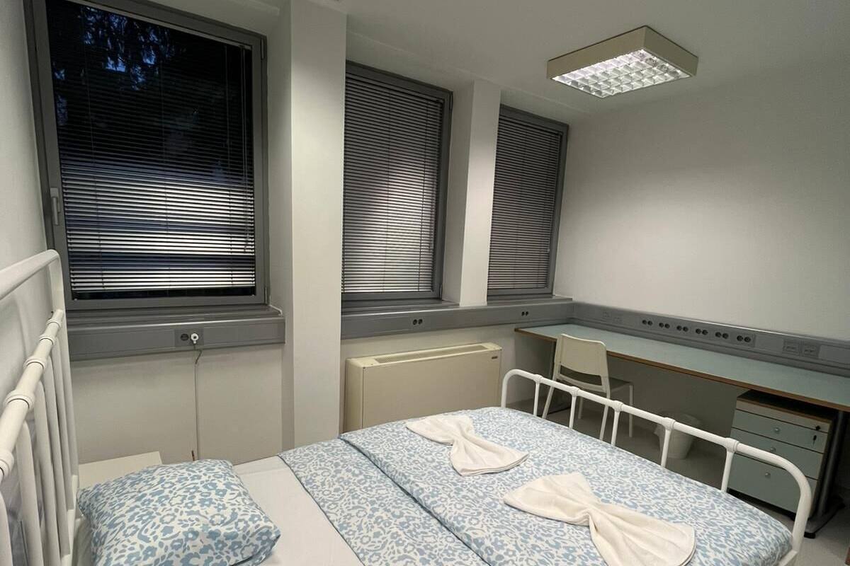 Quarto com cama, mesas de cabeceira, escrivaninha e janela com persianas.