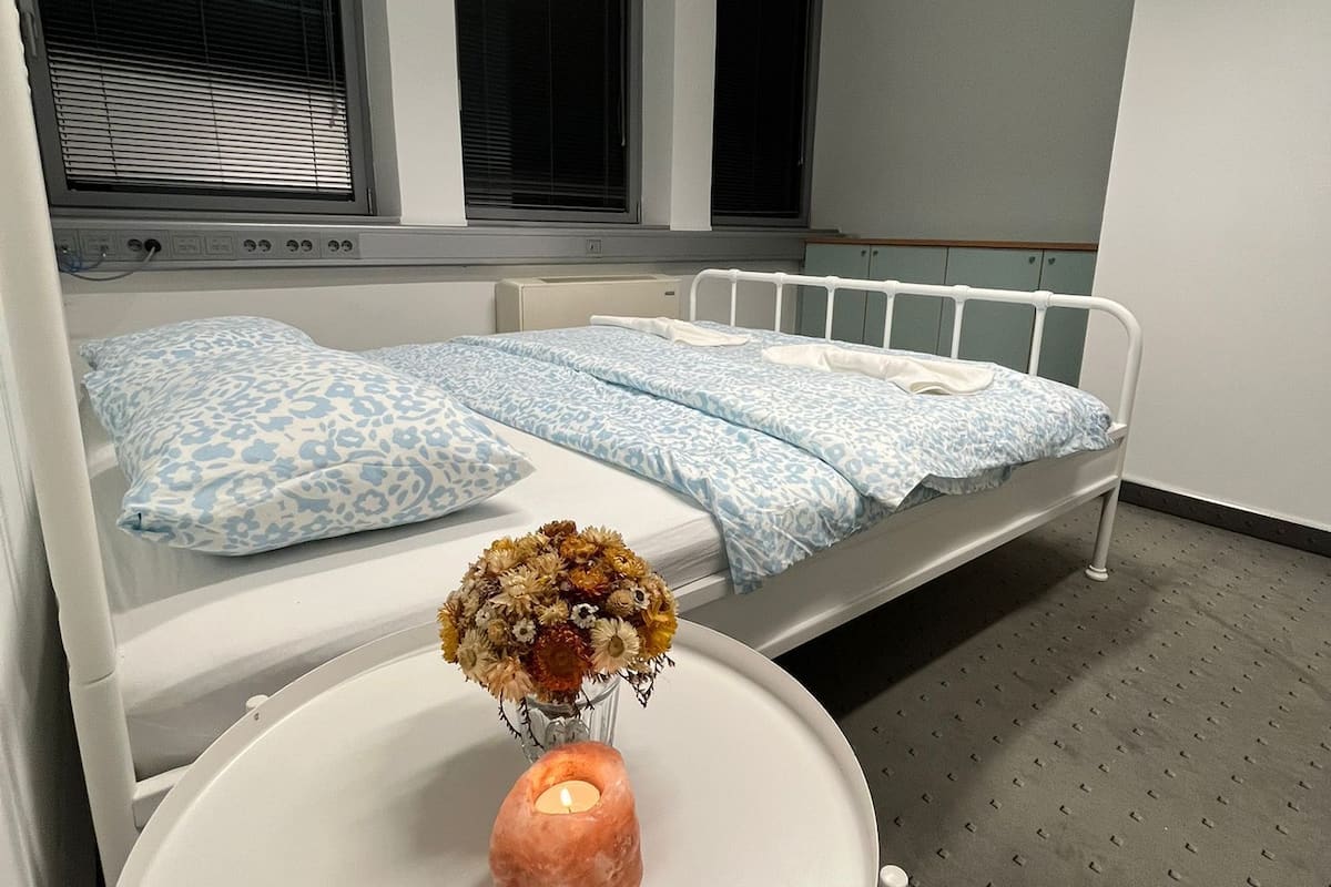 Quarto de hospital com uma cama, uma mesa pequena com um vaso e uma vela, e janelas com persianas.