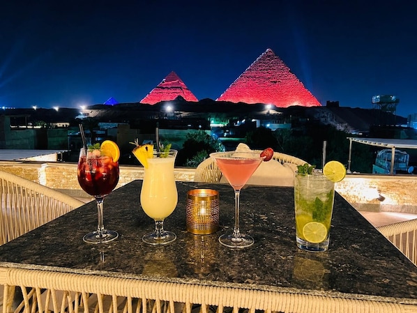 Uma mesa com cinco bebidas, uma vela dourada e duas pirâmides iluminadas ao fundo.