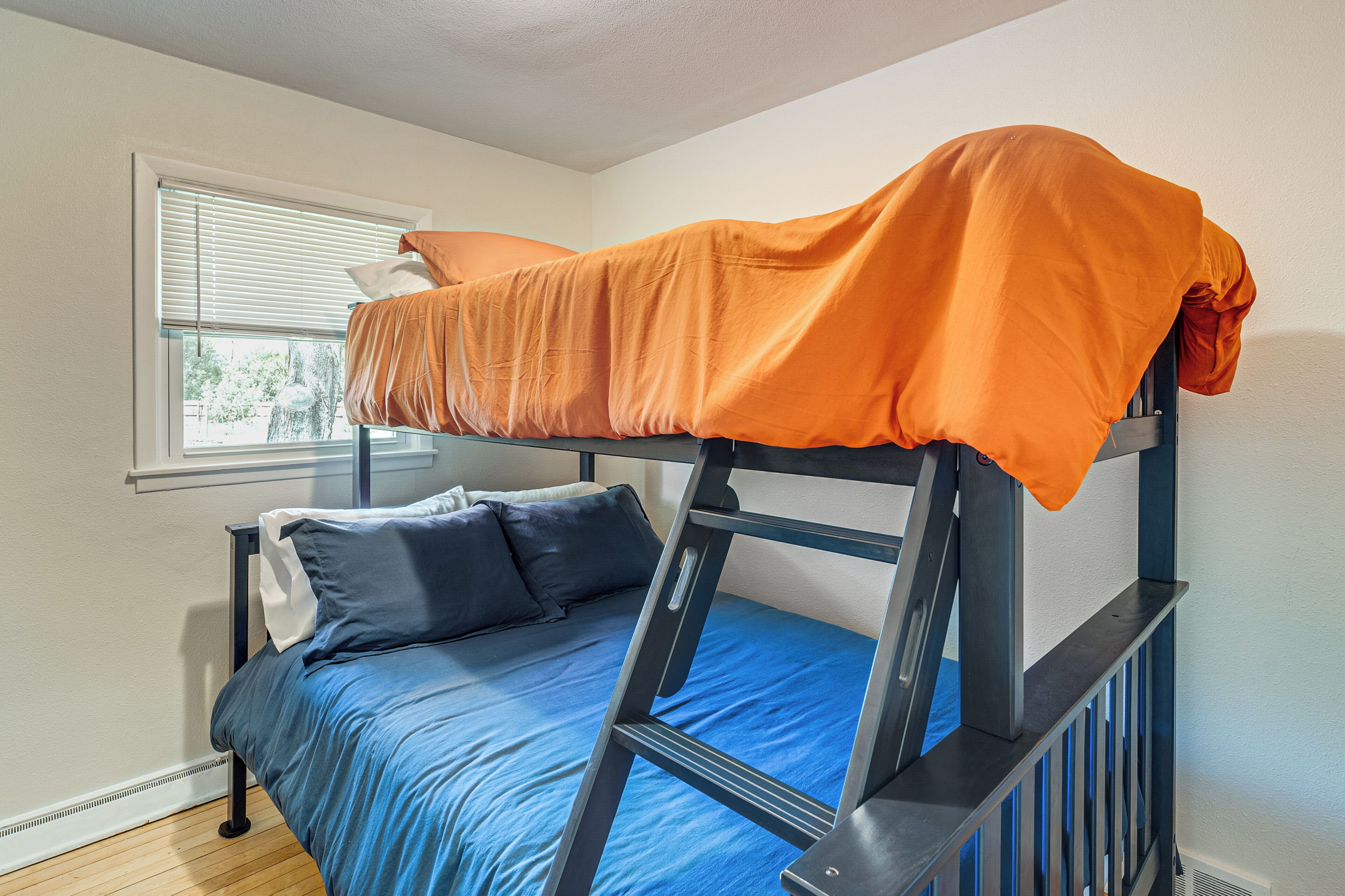 Uma cama beliche com colchão azul e cobertor laranja, uma janela com persianas e uma parede branca.