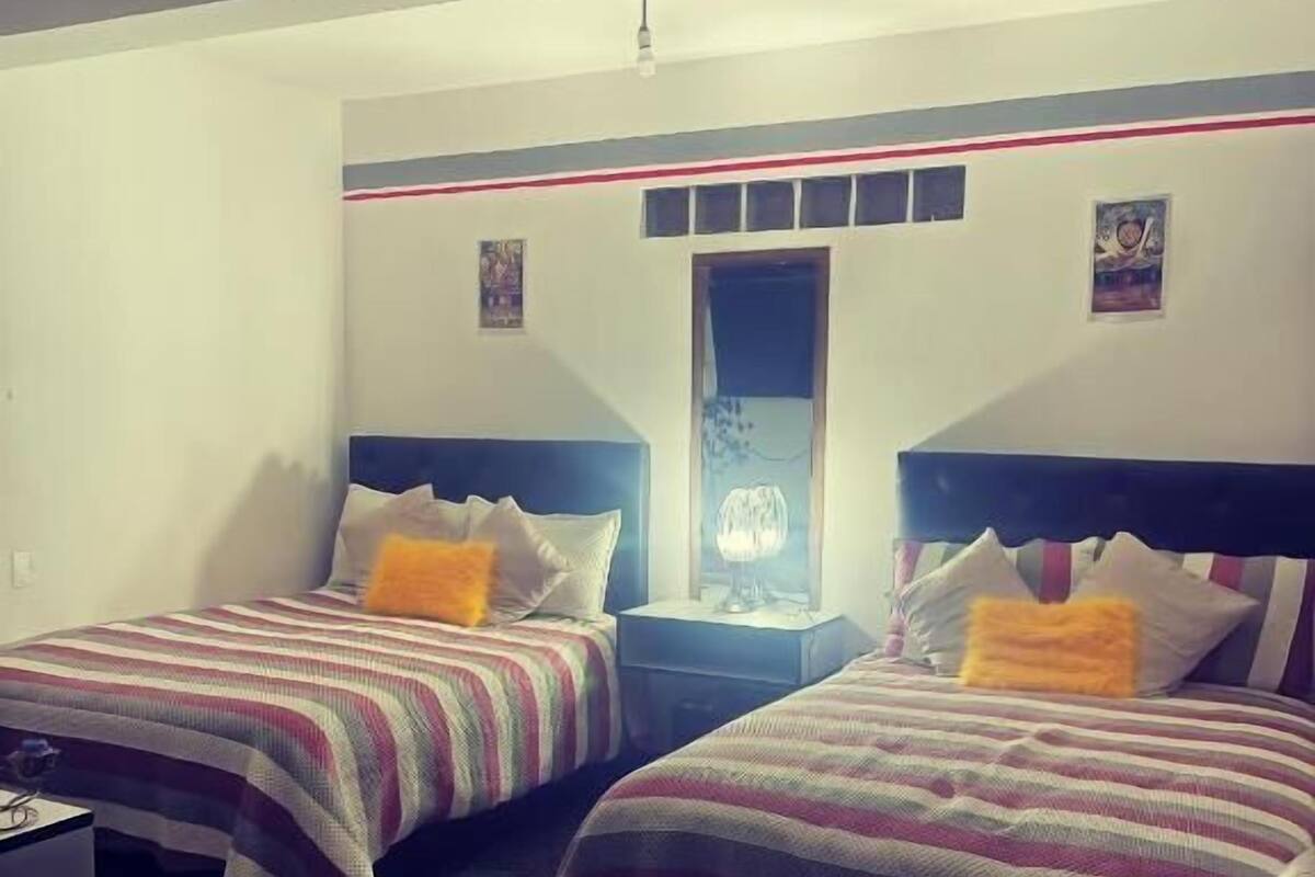 Quarto de hotel com duas camas, uma mesinha, um sofá e uma mesa de cabeceira.
