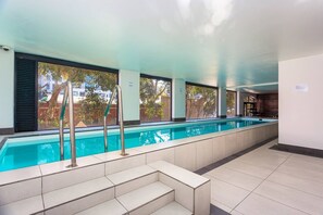 Uma piscina interior com paredes de vidro transparente, degraus de acesso e vista para o exterior.