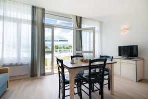 Quarto moderno com mesa de jantar, cadeiras, TV e vista para um edifício através de grandes janelas.