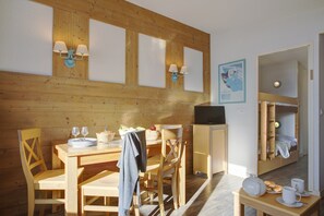 Interior de madeira com uma mesa de jantar, cadeiras, uma mesinha com xícaras e um bule, e uma cama beliche com uma televisão.