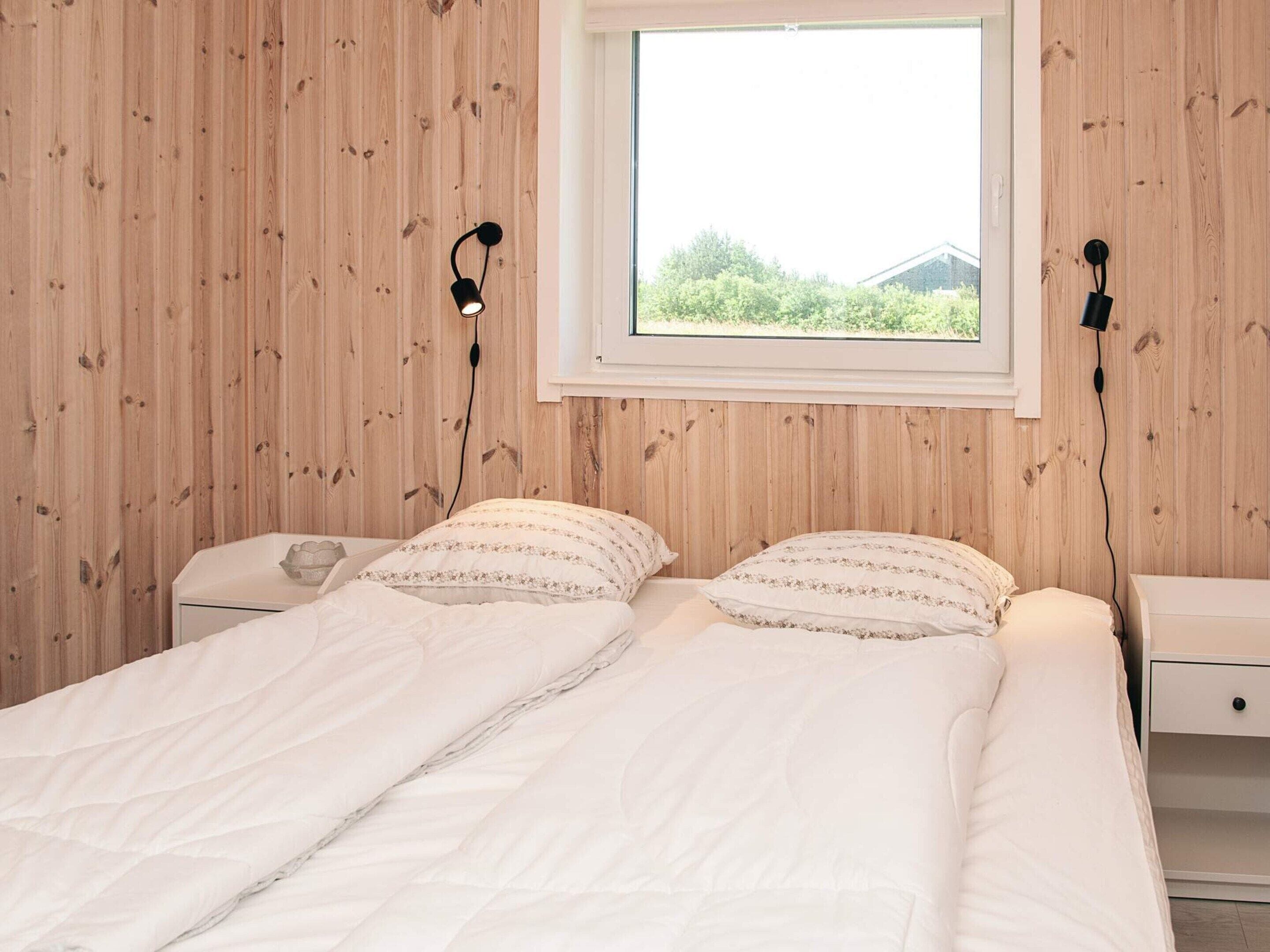 Quarto com parede de madeira, janela, cama com roupa de cama branca e abajures laterais.