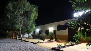 Um edifício com uma placa indicando 'Los Palmitos' à noite, cercado por cascalho, árvores e pequenas plantas.
