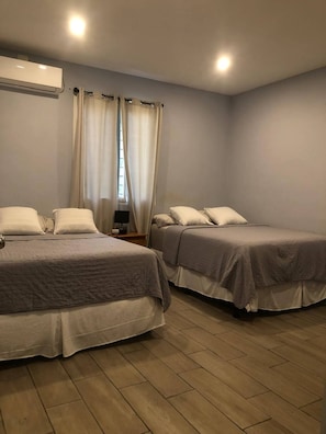 Quarto com duas camas, janela com cortinas, ar-condicionado e piso de madeira.