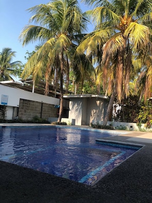 Piscina rodeada por palmeiras, um edifício de concreto e céu claro.