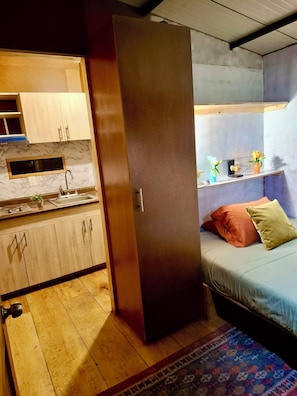 Quarto compacto com cozinha americana, cama com almofadas e tapete estampado.