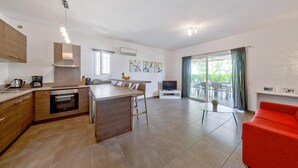 Uma cozinha moderna com uma ilha central, uma sala de jantar com mesa e cadeiras, uma sala de estar com sofá vermelho e uma porta de vidro deslizante para o exterior.
