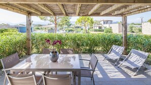 Um alpendre coberto com mesa e cadeiras, rodeado por vegetação e vista para edifícios ao longe.