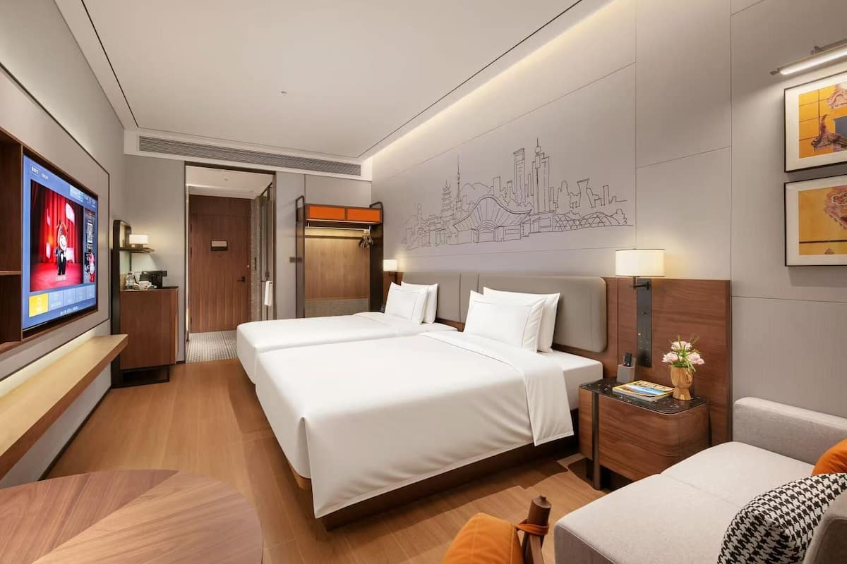 Quarto de hotel com duas camas, televisão, escrivaninha, cadeira e parede com um mural da paisagem urbana.