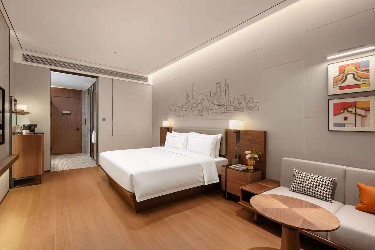 Quarto de hotel com cama, sofá, pequena mesa, mesa de cabeceira e arte de parede com vista para a cidade.