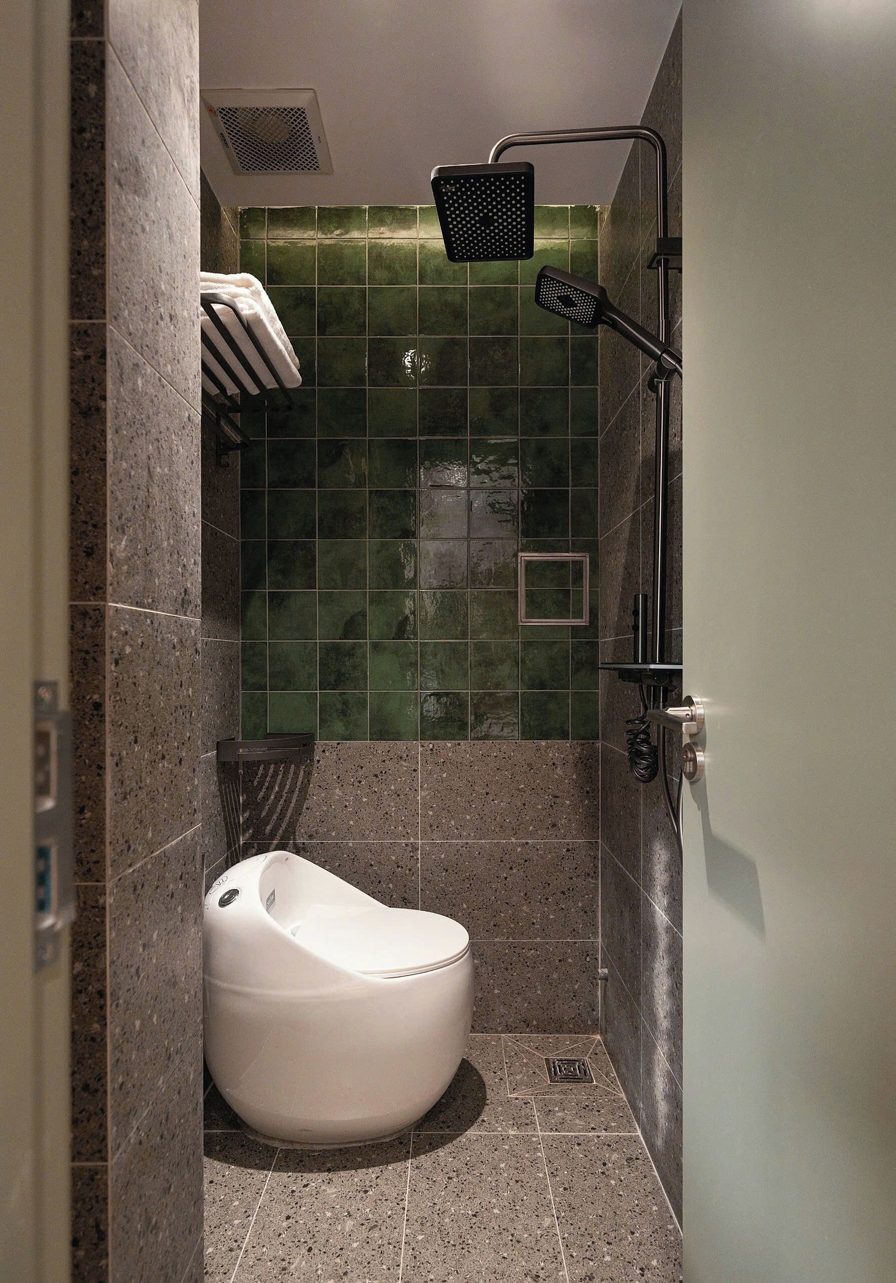 Um banheiro moderno com vaso sanitário branco, chuveiro com haste móvel e paredes revestidas de azulejos verdes.