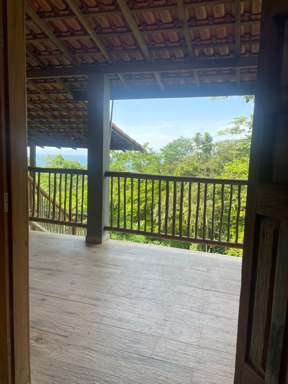 Um terraço de madeira aberto com vista para vegetação exuberante e um prédio ao longe.
