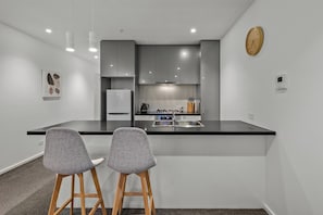 Cozinha moderna com bancada preta, eletrodomésticos de aço inoxidável e dois bancos altos cinzentos.