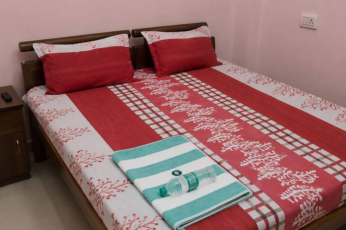 Uma cama bem arrumada com um cobre-leito estampado em vermelho e branco, duas almofadas vermelhas e uma toalha dobrada com uma faixa verde.