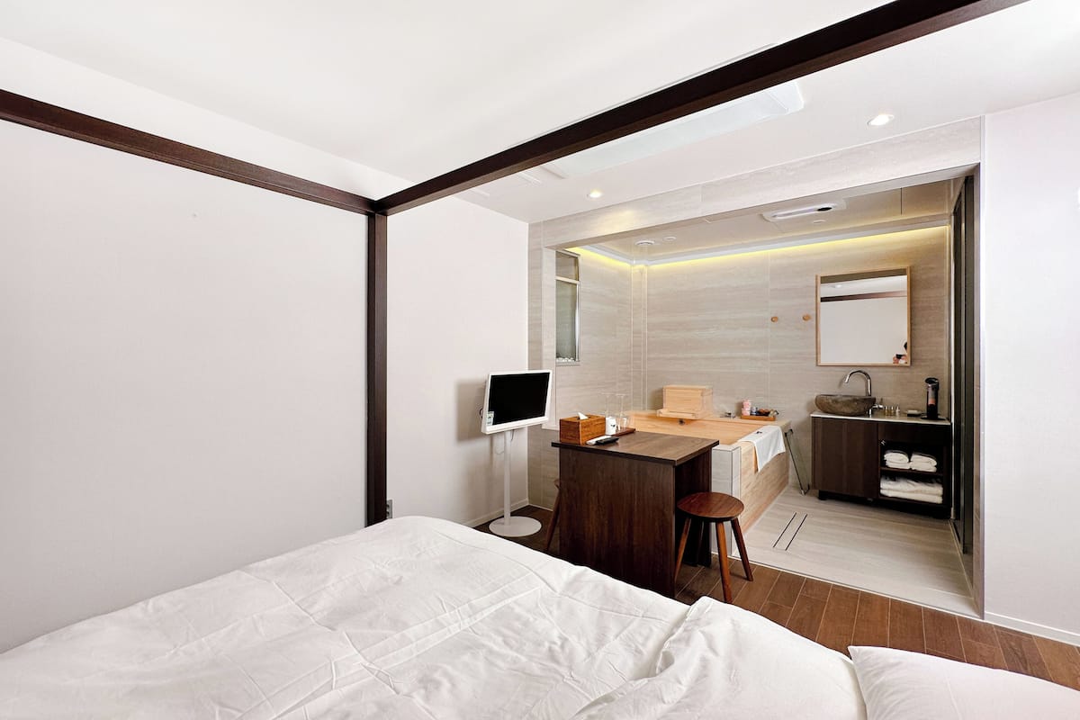 Quarto de hotel moderno com cama, escrivaninha, espelho e banheiro visível por uma porta aberta.