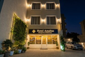 Entrada de hotel com uma placa indicando 'Hotel Amaltas Managed By OTABootup', plantas em vasos e um carro estacionado na frente.