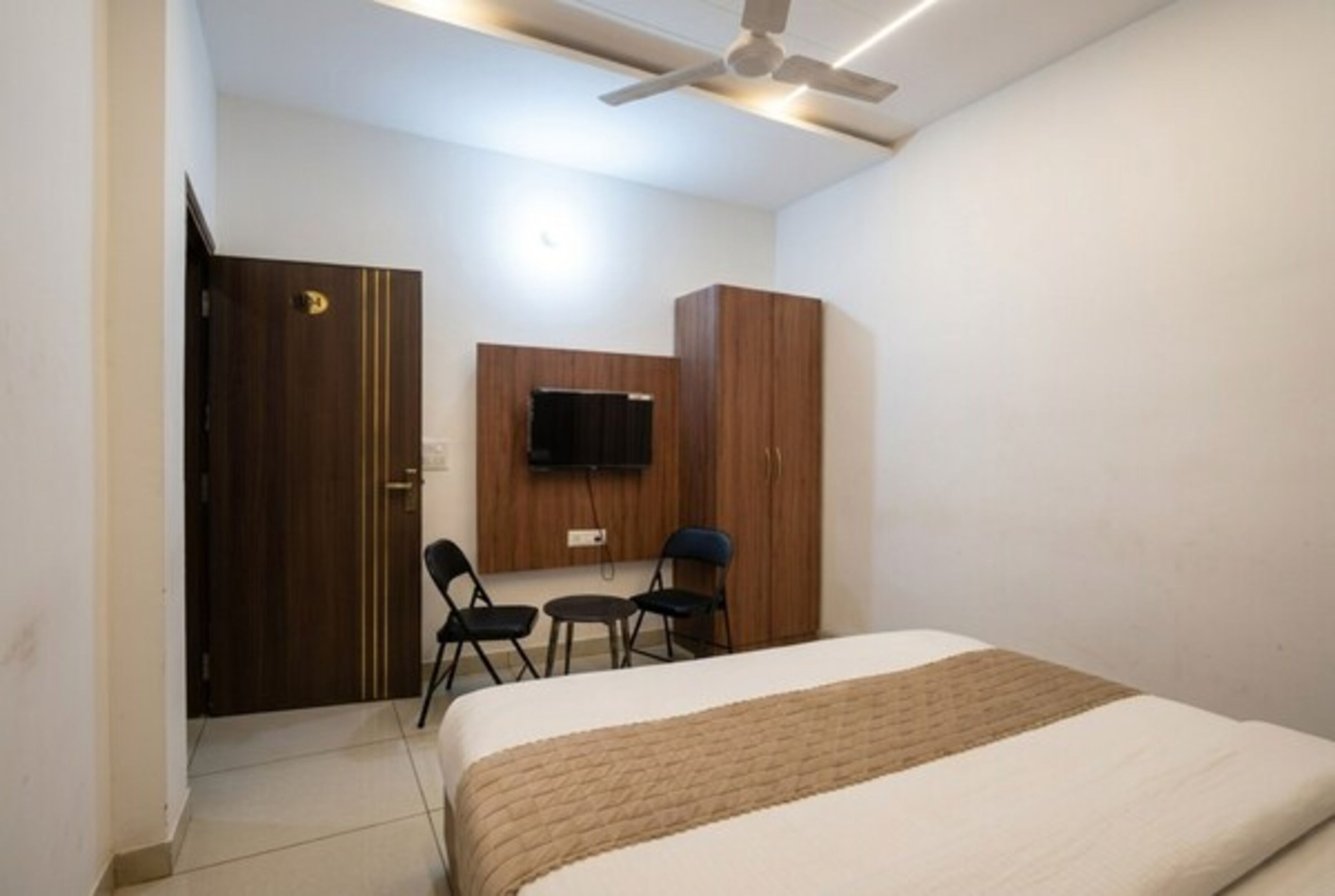 Quarto com cama, escrivaninha, duas cadeiras, guarda-roupa, televisão e ventoinha no teto.