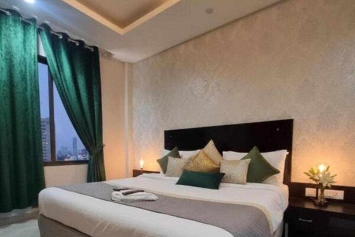 Quarto moderno com uma cama grande, uma mesinha de cabeceira, e vista da cidade através de uma janela com cortinas verdes.