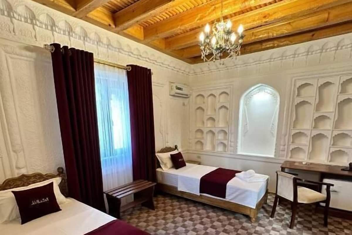 Quarto de hotel com piso xadrez, duas camas, uma escrivaninha e um lustre.