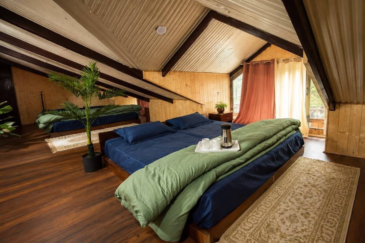 Um quarto com teto inclinado, paredes de madeira, uma cama grande com roupa de cama azul e verde, um vaso de planta e uma janela com cortinas.