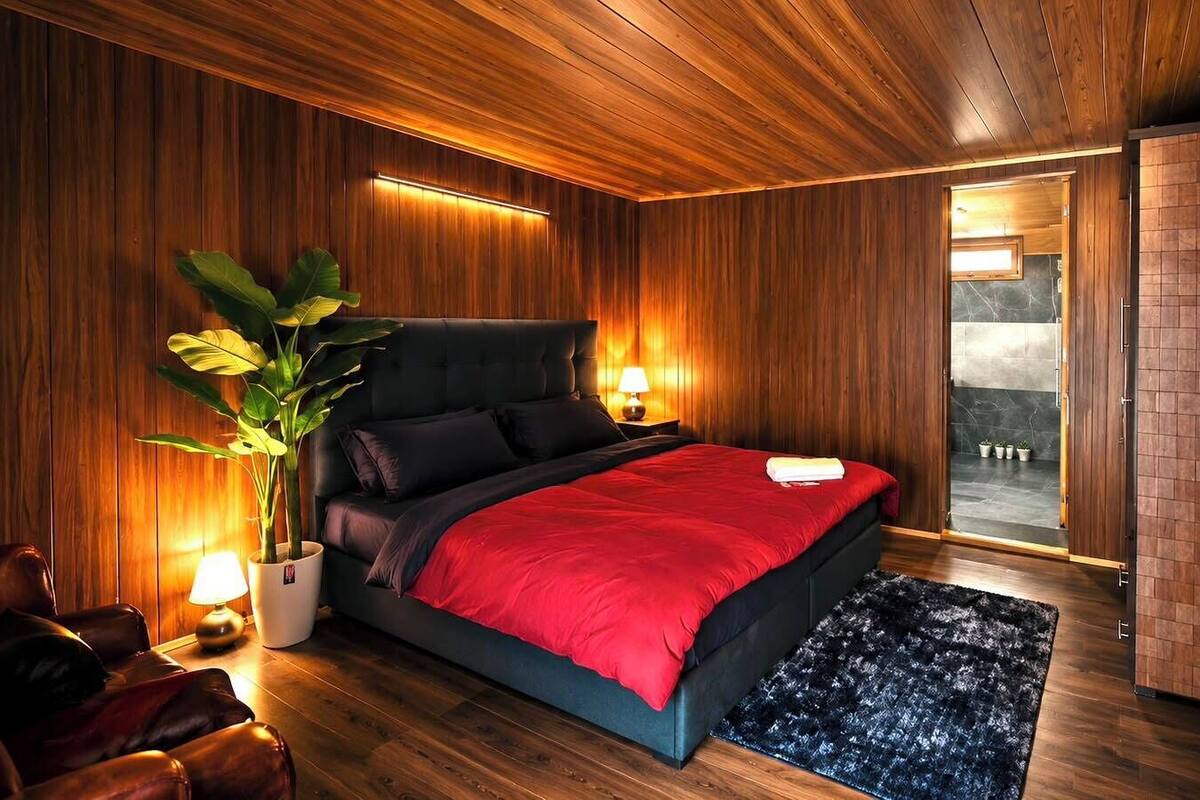 Quarto com cama grande, paredes de madeira, planta em vaso e vista através de uma porta aberta.