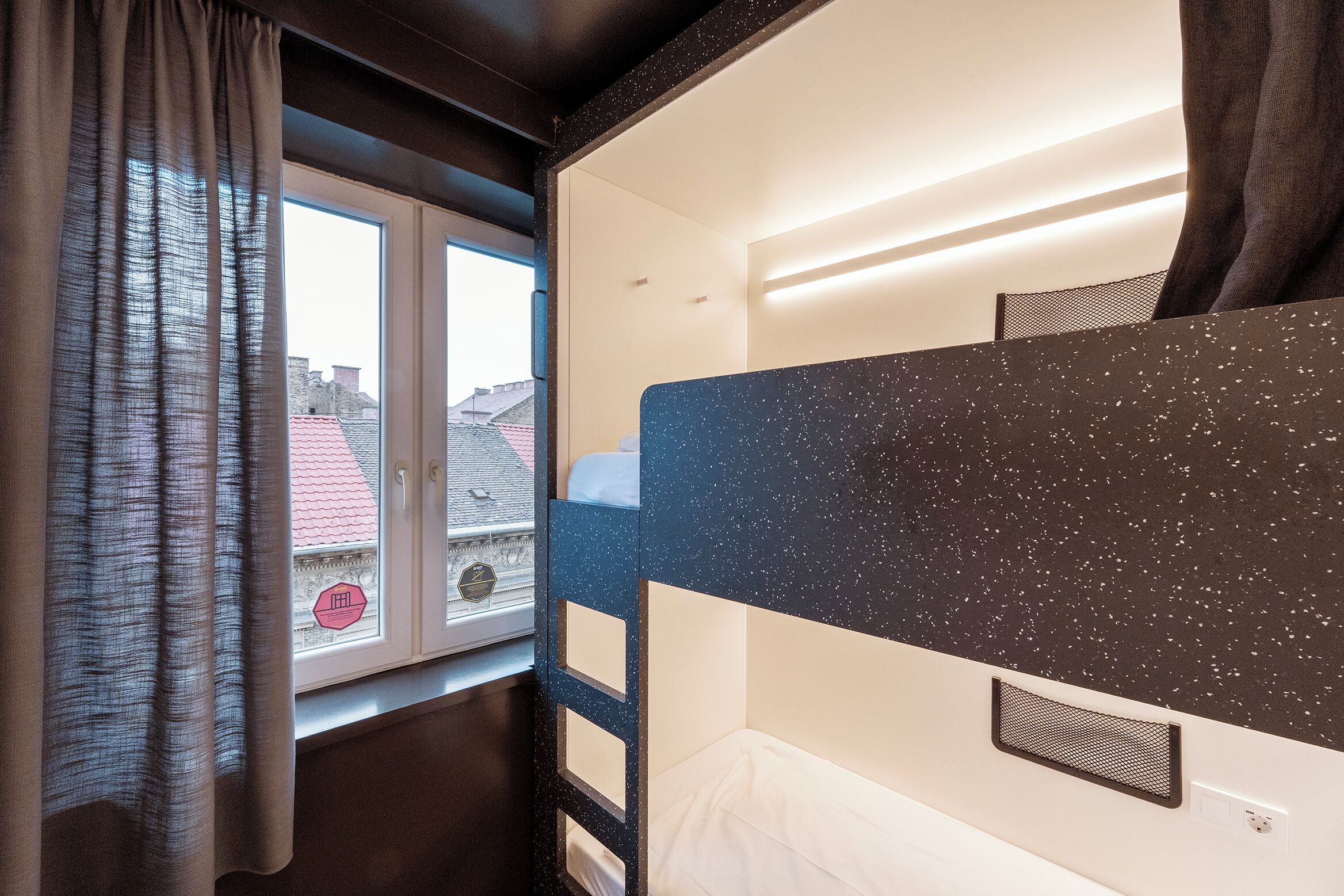 Uma cama de solteiro com um desenho de estrelas, uma janela com vista para prédios e uma cortina.