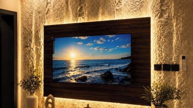 Televisão fixada na parede com vista para o pôr do sol à beira-mar, um console de madeira abaixo e parede de pedra texturizada.