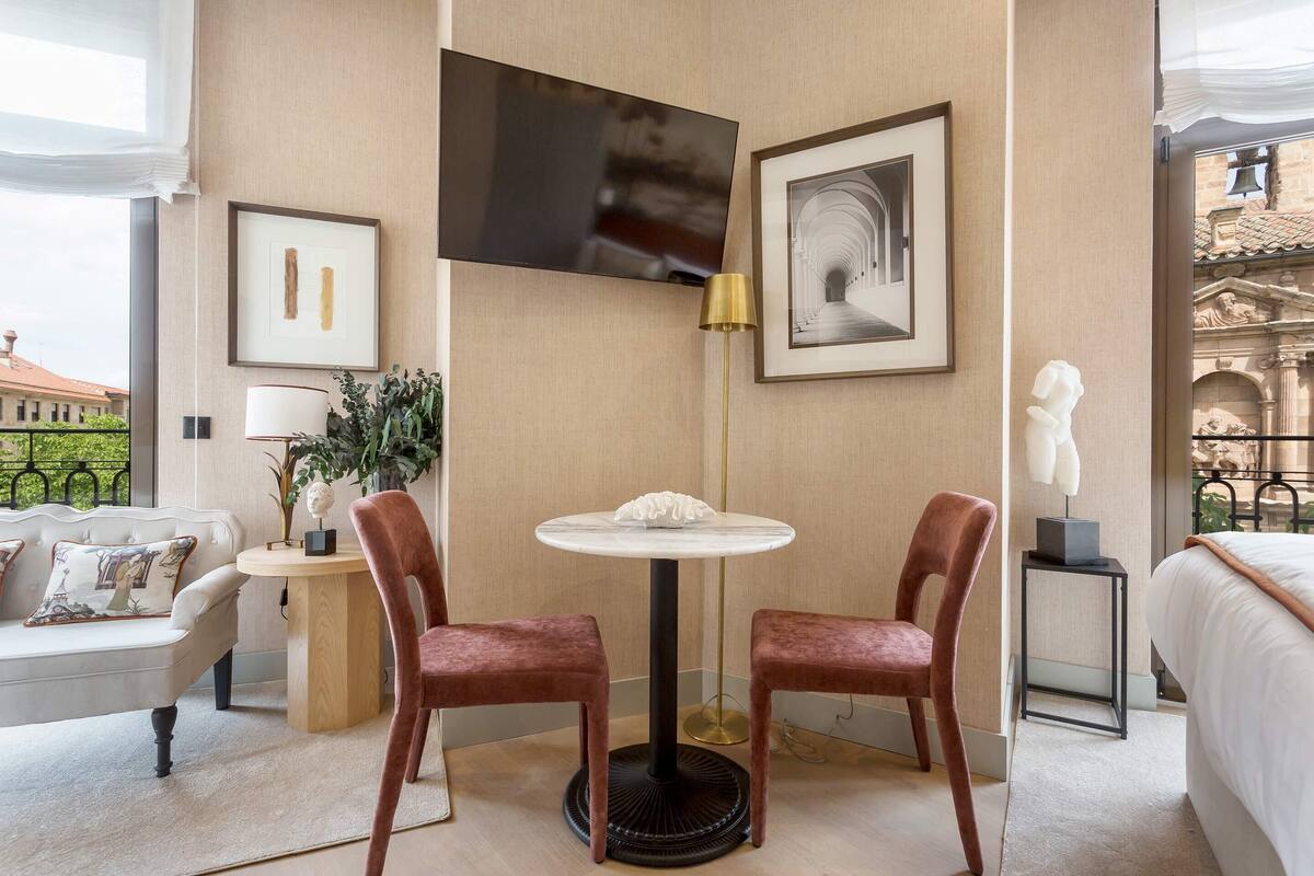 Quarto de hotel com uma mesa redonda, duas cadeiras, um sofá, uma televisão, obras de arte emolduradas, um abajur e uma estátua.