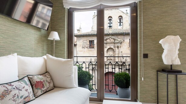 Quarto de hotel moderno com sofá, televisão de tela plana, varanda com plantas em vasos e vista para edifícios históricos.
